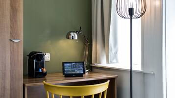 Um quarto com escrivaninha de madeira, uma cadeira amarela, máquina de café, laptop, luminária e janela com cortinas.