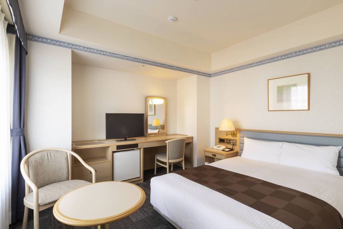 Quarto de hotel com cama, escrivaninha, cadeira, televisão, mesa de cabeceira, abajur e um quadro na parede.
