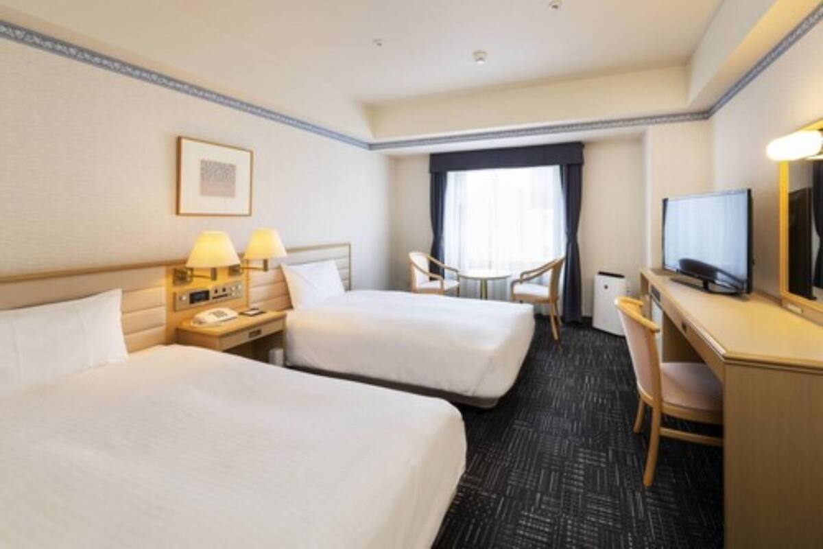 Quarto de hotel com duas camas, uma escrivaninha com televisão, uma cadeira e uma mesinha.