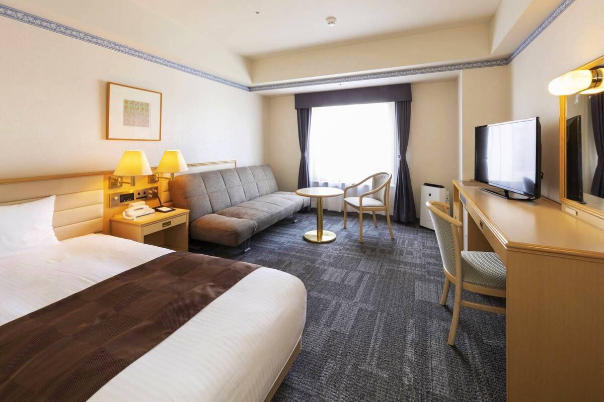 Quarto de hotel com cama, sofá, escrivaninha com televisão e uma cadeira.