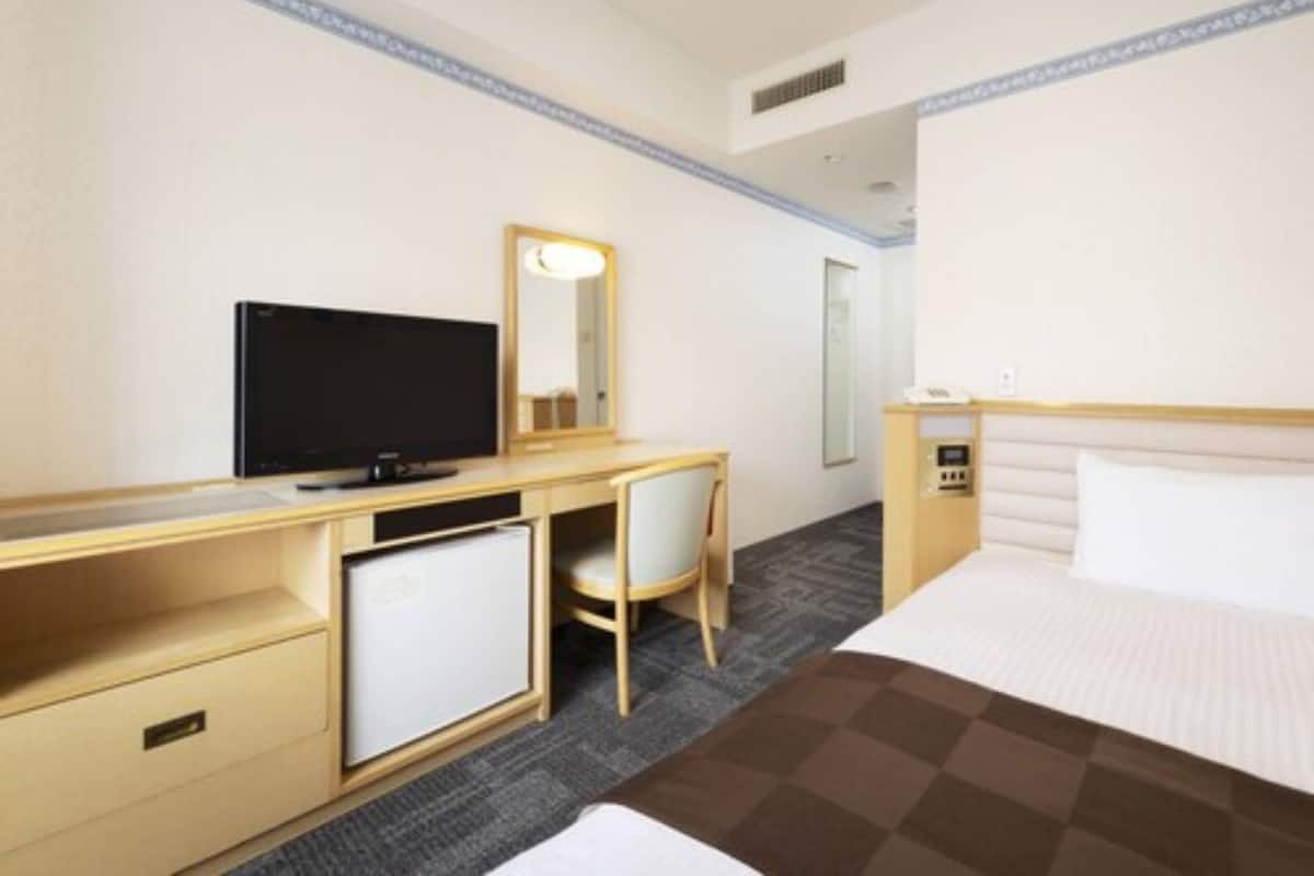 Quarto de hotel com uma cama, uma escrivaninha com televisão, uma cadeira e um pequeno frigorífico.