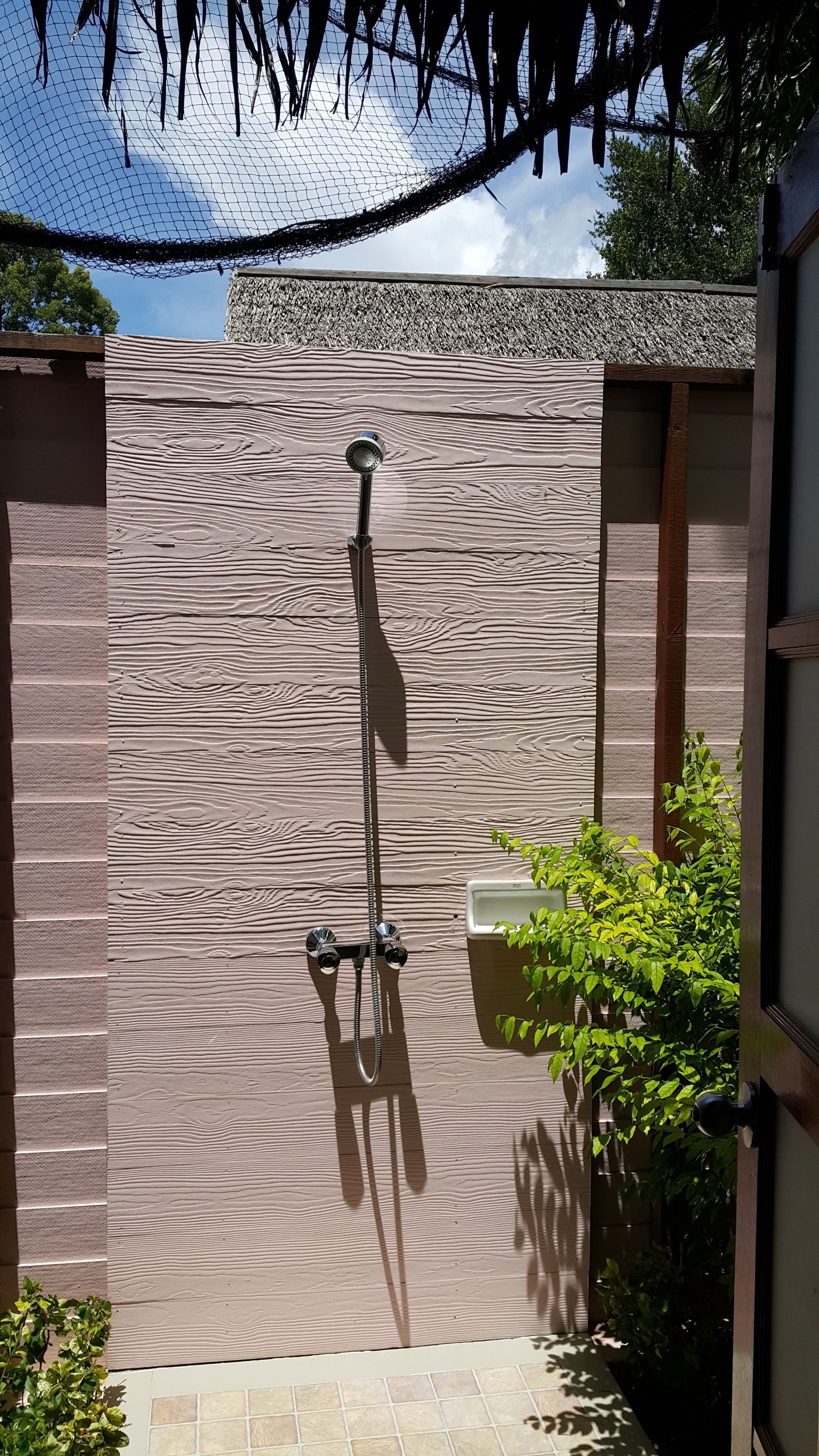 Um chuveiro externo moderno com parede de textura amadeirada, mangueira de metal e recipiente branco.