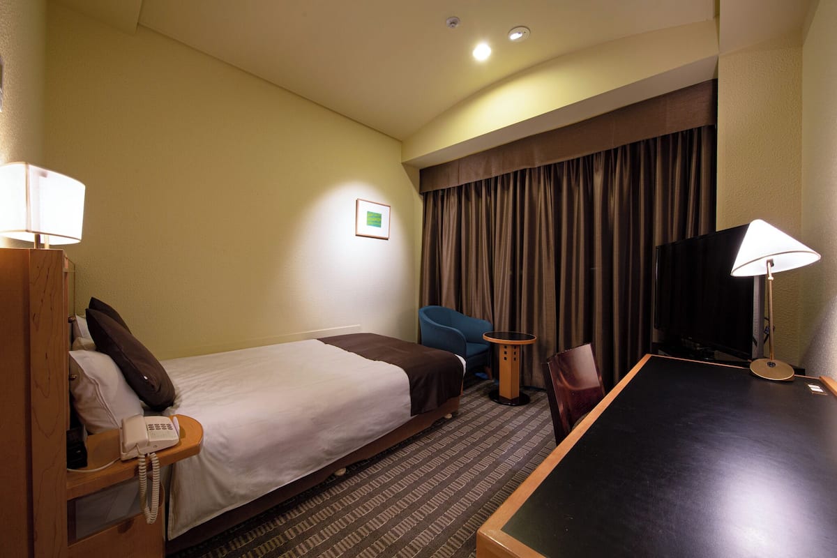 Quarto de hotel com cama, escrivaninha, cadeira, televisão e telefone.