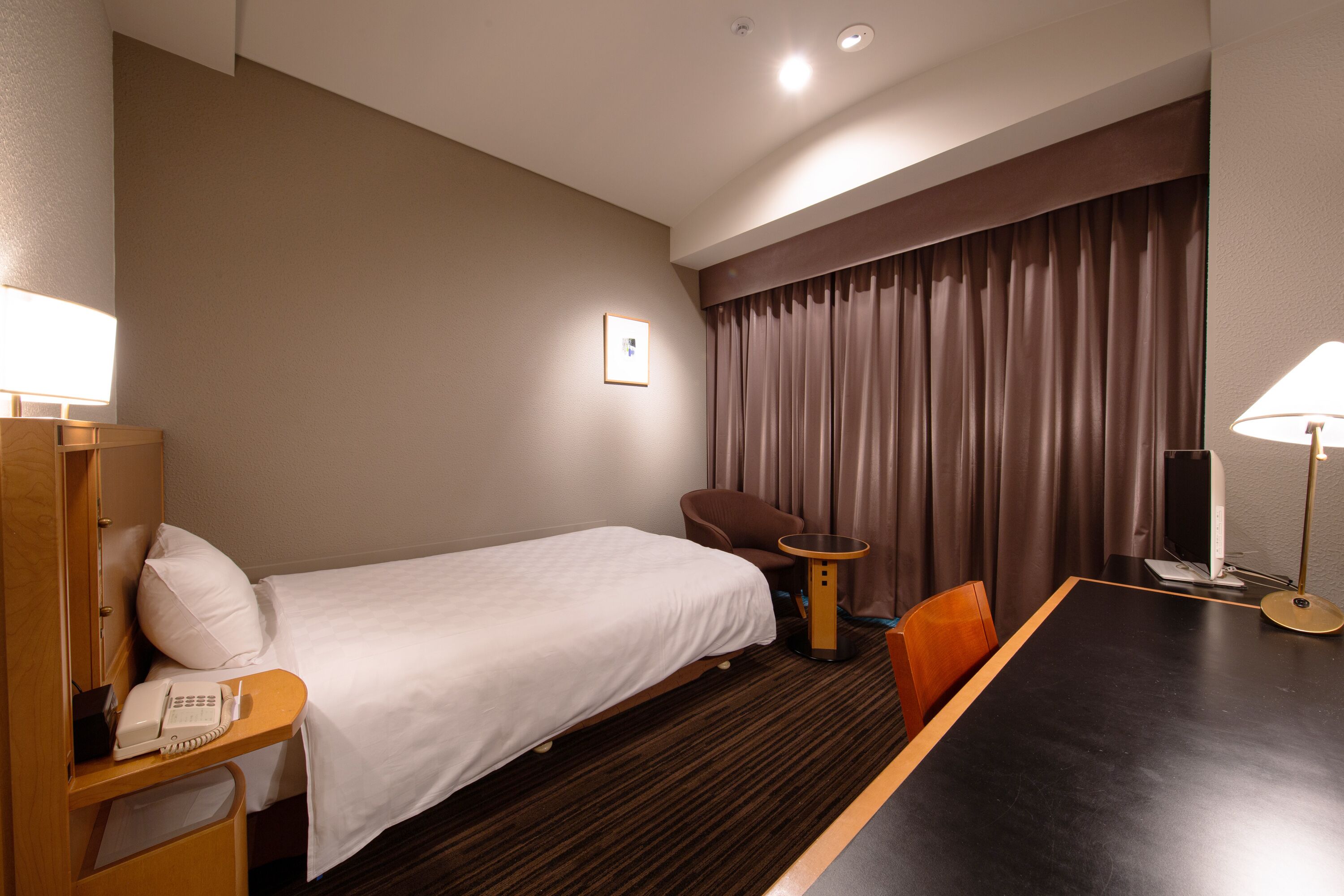 Quarto de hotel com uma cama, uma escrivaninha com candeeiro, uma cadeira, uma mesinha e uma cortina.