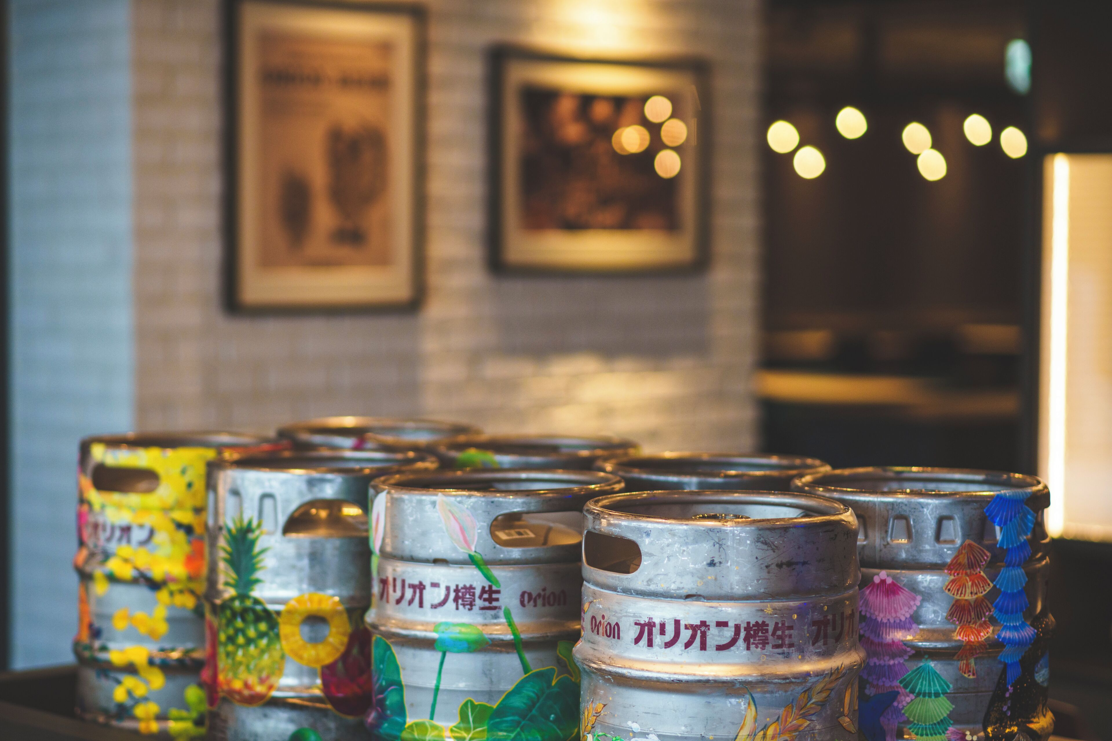 Dispensadores de bebidas em metal com etiquetas coloridas, parede de tijolos e quadros emoldurados ao fundo.