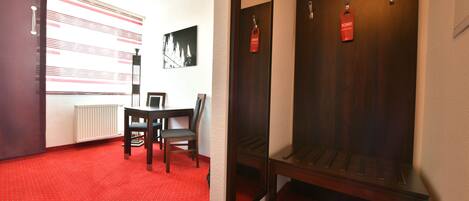 Quarto de hotel com tapete vermelho, um banco de madeira, mesa com cadeiras, radiador e um quadro fixado na parede.