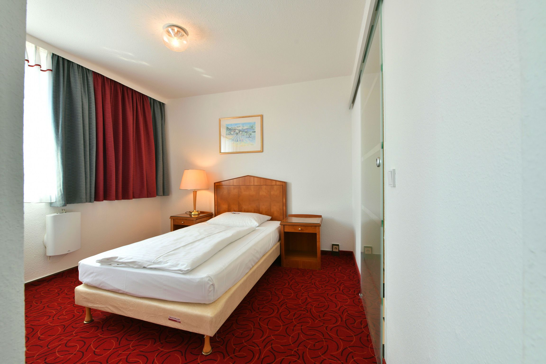 Quarto de hotel com uma cama de solteiro, mesa de cabeceira com abajur, cabeceira de madeira, janela com cortinas e um quadro na parede.
