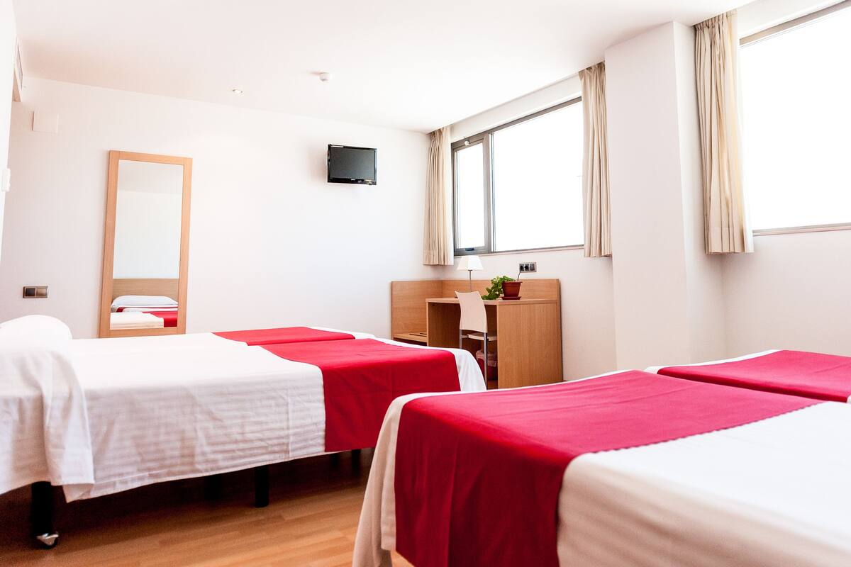 Quarto de hotel com duas camas, uma escrivaninha, um espelho, uma televisão e uma janela com cortinas.