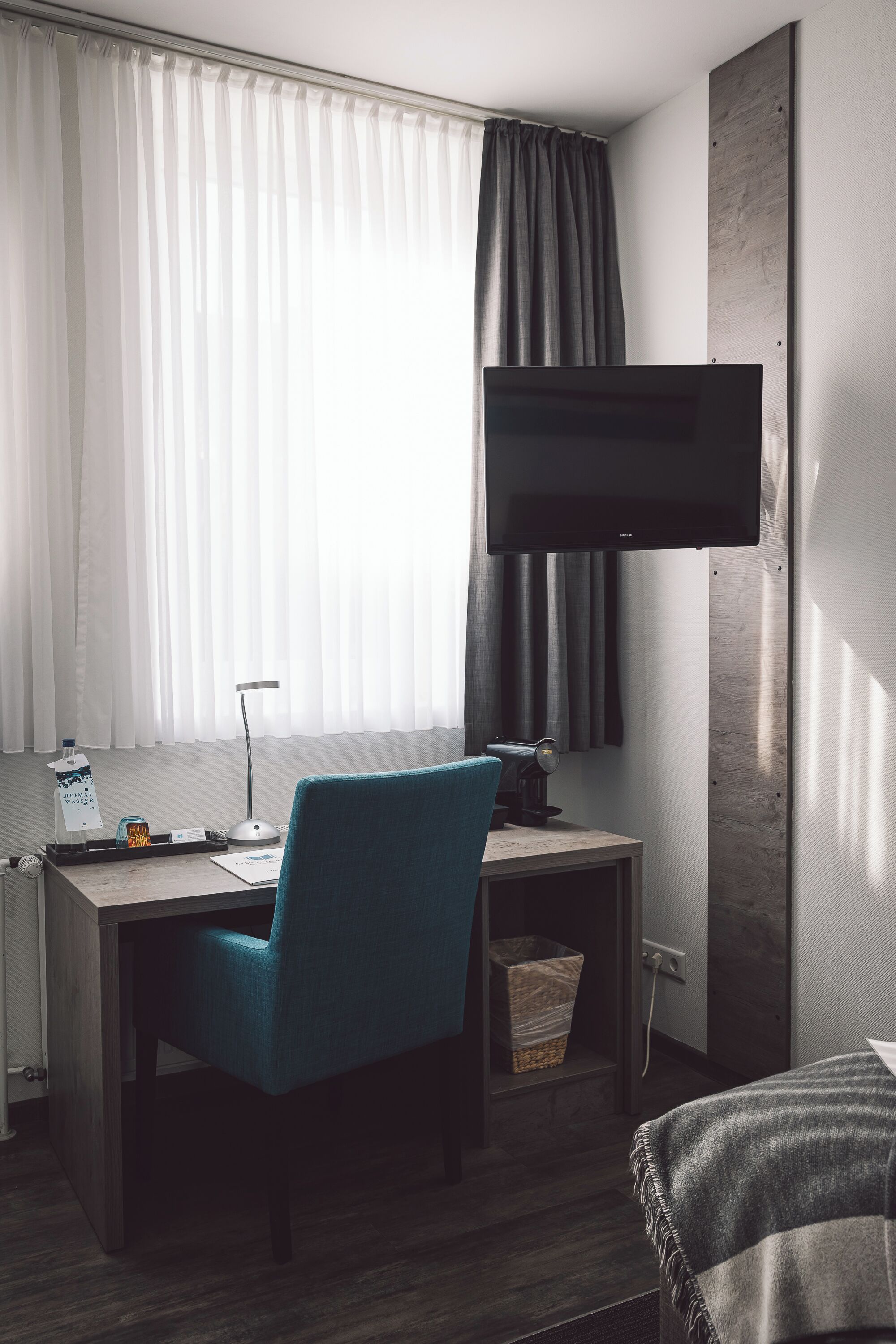Quarto de hotel moderno com escrivaninha, cadeira, televisão e janela com cortinas.
