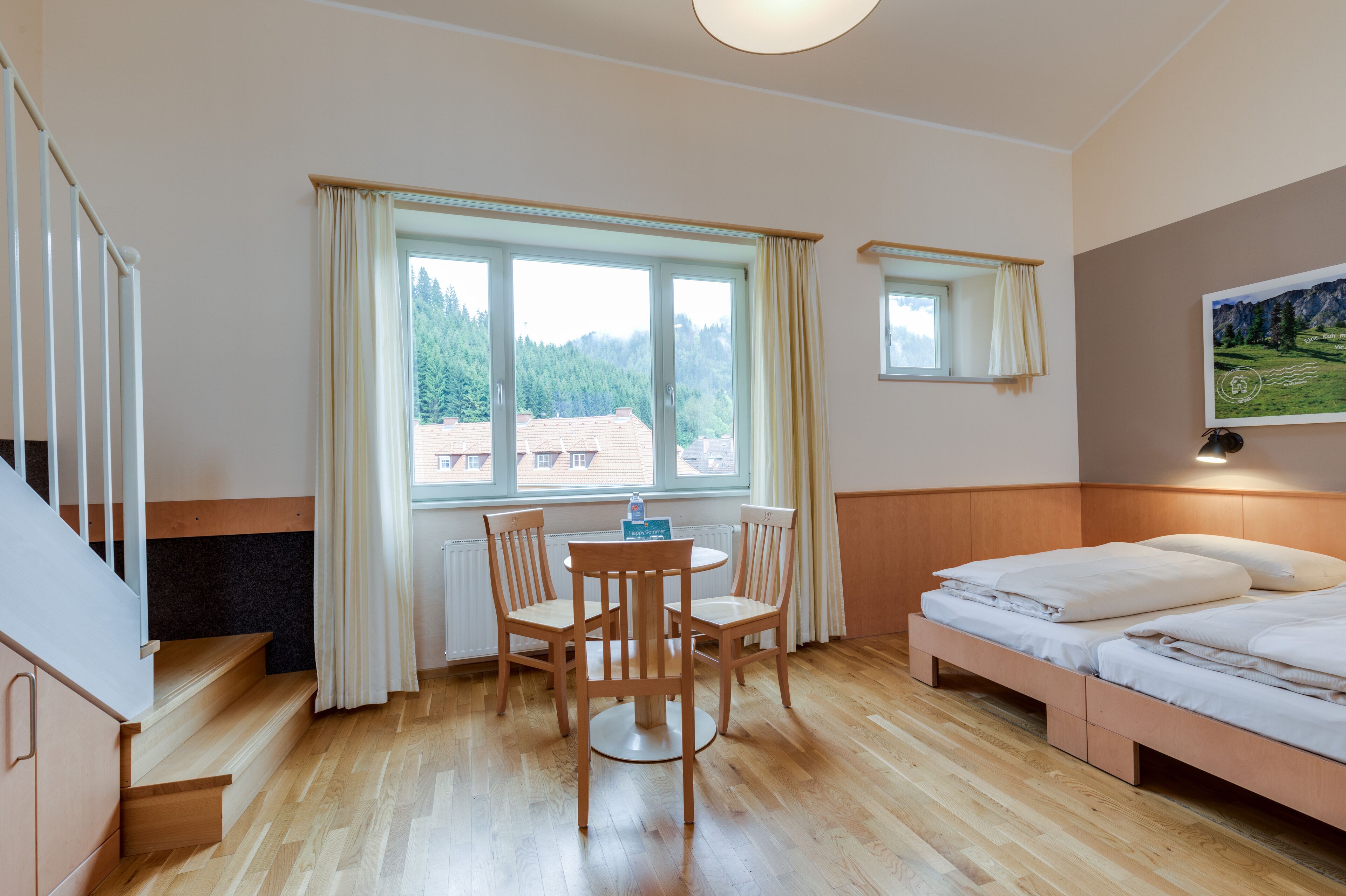 Quarto com cama, mesa com cadeiras, janela com cortinas e um quadro na parede.