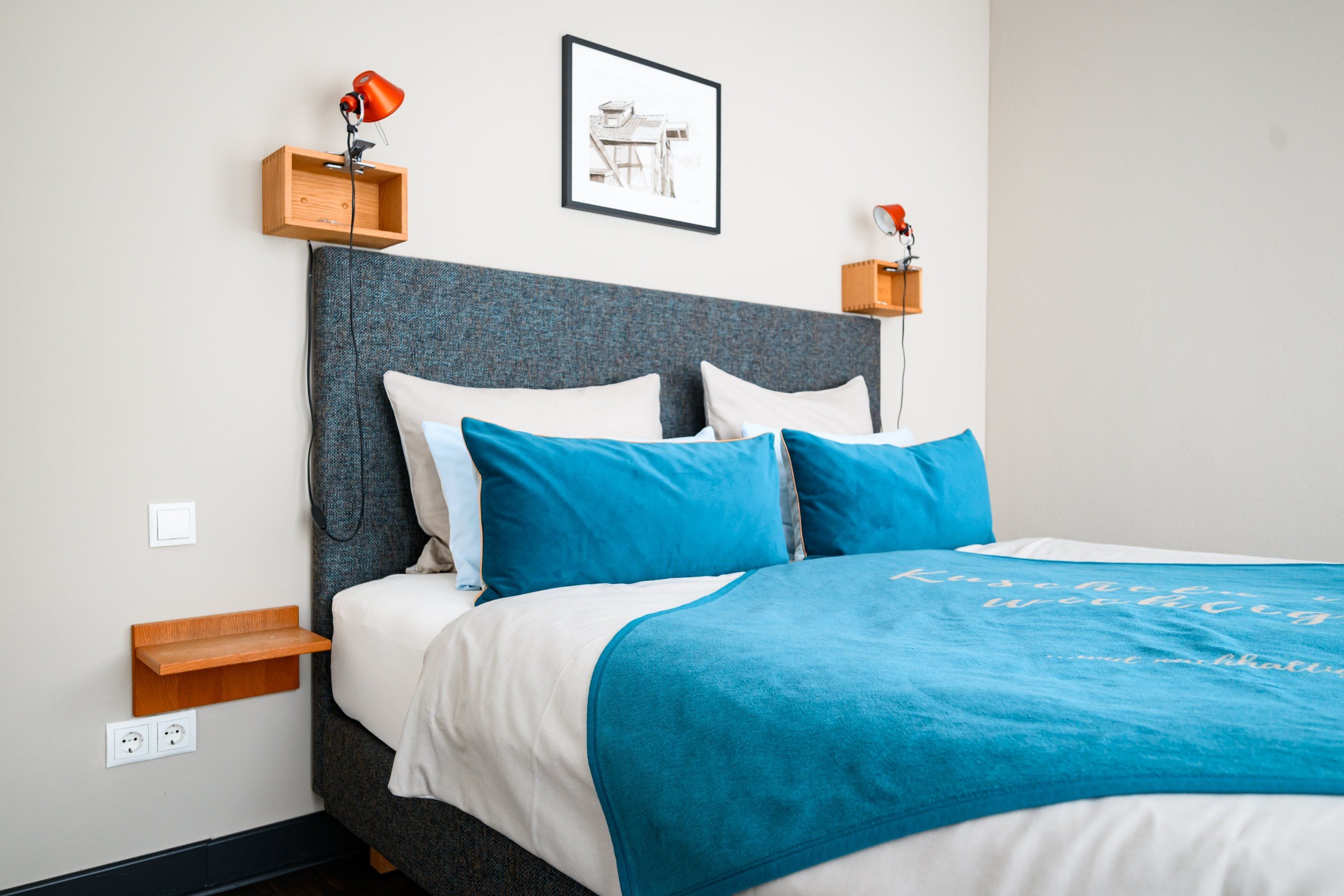 Cama bem arrumada com edredom azul e branco, dois travesseiros e um cobertor azul. Há duas mesas de cabeceira com luminárias, um quadro na parede e uma pequena estante de madeira.