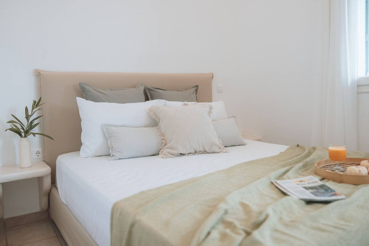 Uma cama bem arrumada com cabeceira bege, roupa de cama branca e vários travesseiros. Uma mesa de cabeceira com uma planta e um abajur são visíveis.