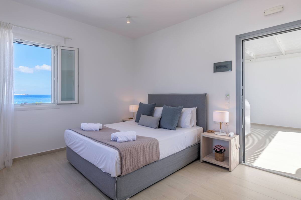 Um quarto com cama, mesas de cabeceira, cortinas e vista para o mar.