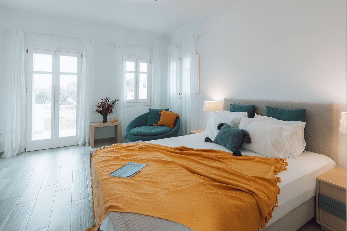 Quarto com cama, mesa de cabeceira, cadeira e janela com cortinas.