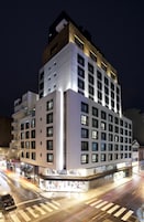 Um hotel de vários andares com design moderno, em uma localização de destaque na esquina, próximo a uma faixa de pedestres e lojas.