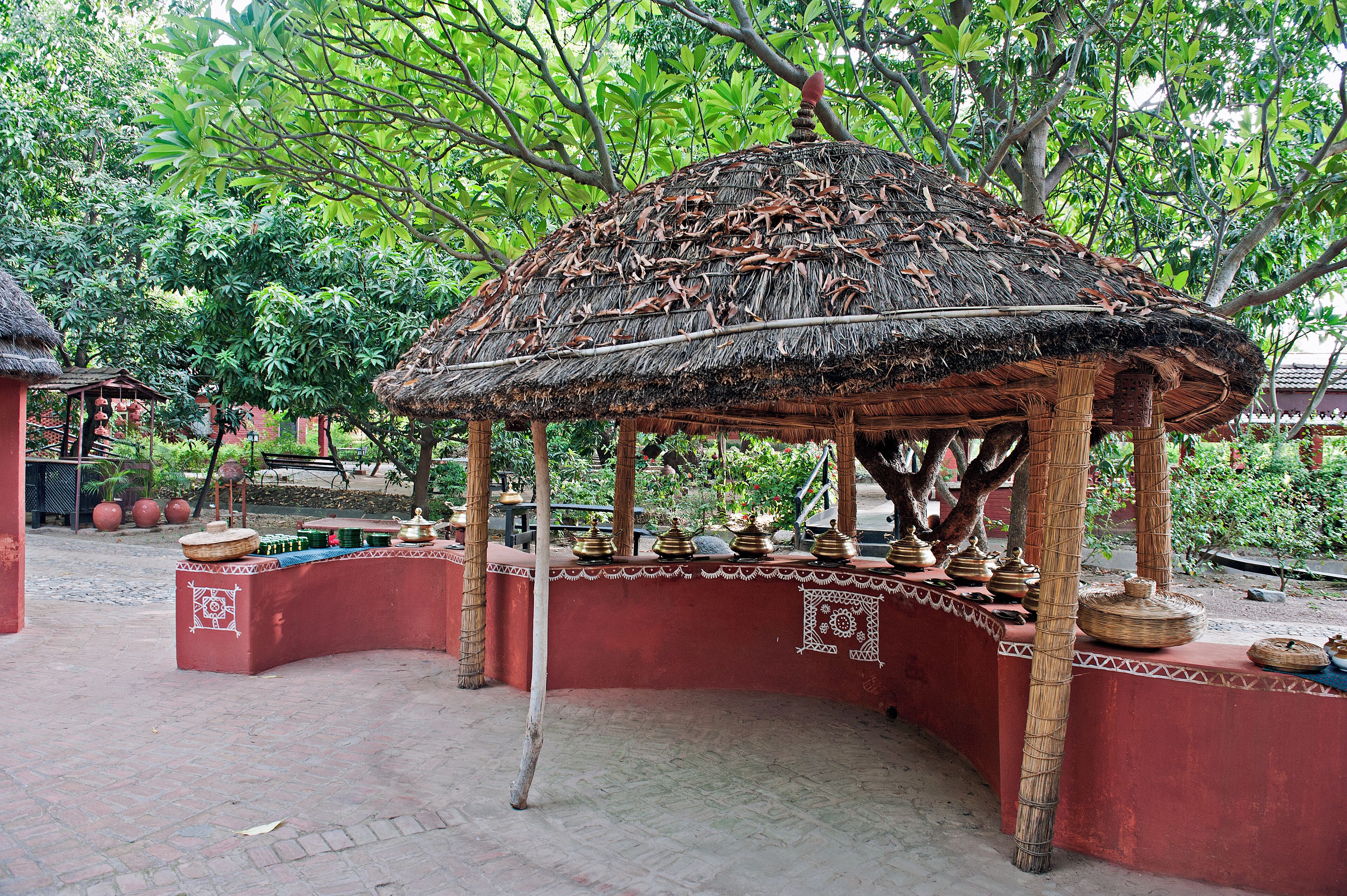 Um pavilhão tradicional com telhado de palha e parede curva vermelha, rodeado por vegetação exuberante.