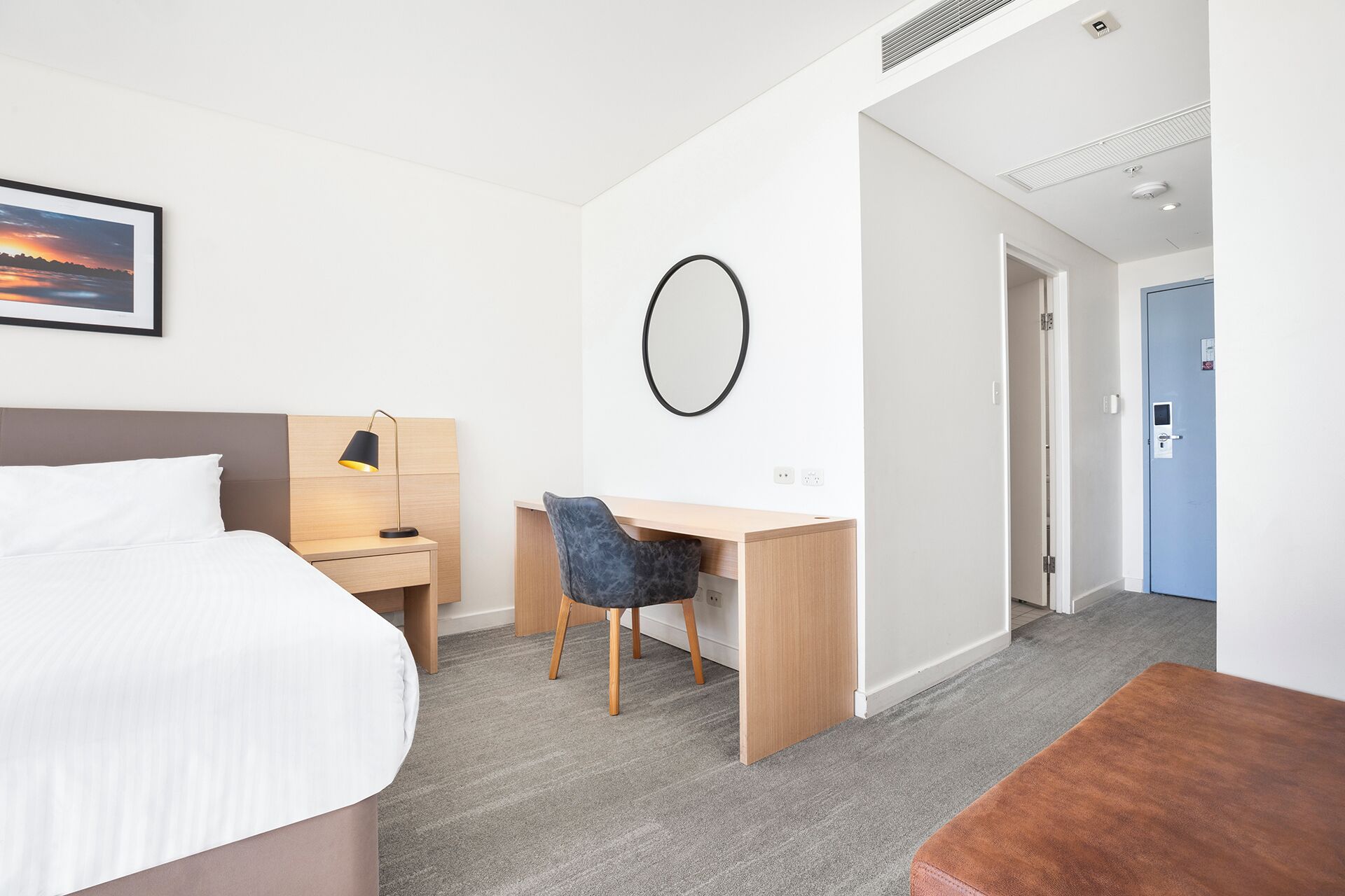Quarto de hotel com cama, escrivaninha, cadeira, espelho redondo e porta.