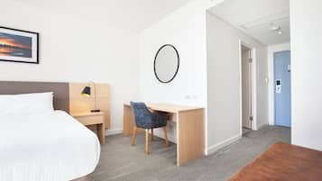 Quarto de hotel com cama, escrivaninha, cadeira, espelho redondo e porta.