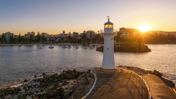 Um farol junto a um porto calmo, com barcos, céu claro e edifícios ao fundo.