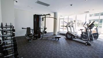 Uma academia com diversos equipamentos de ginástica, amplas janelas e piso coberto por carpete.