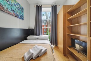Quarto de hotel com uma cama, uma estante de madeira, um quadro na parede e uma janela com cortinas.