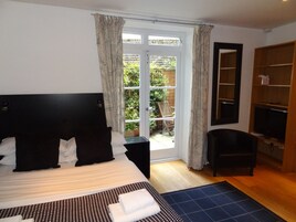 Quarto de hotel com uma cama grande, televisão, pequena escrivaninha, cadeira e vista para um jardim através de uma porta de vidro.