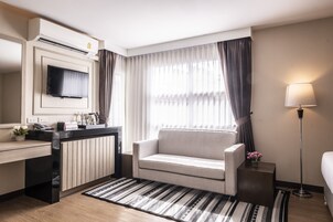 Quarto de hotel com sofá, escrivaninha, televisão de tela plana, abajur e uma janela grande com cortinas.