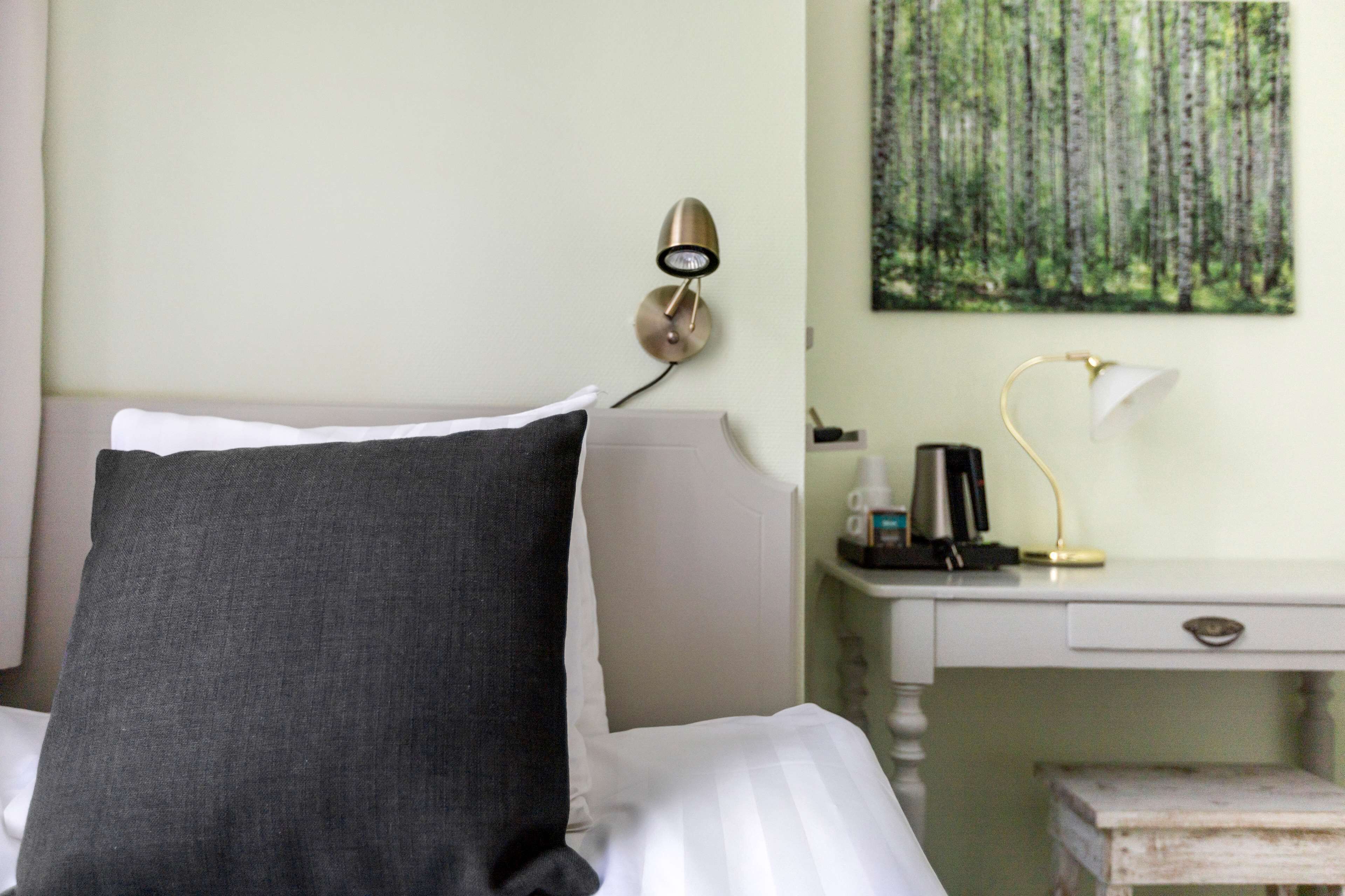 Quarto de hotel com uma cama, escrivaninha, luminária e um quadro na parede.