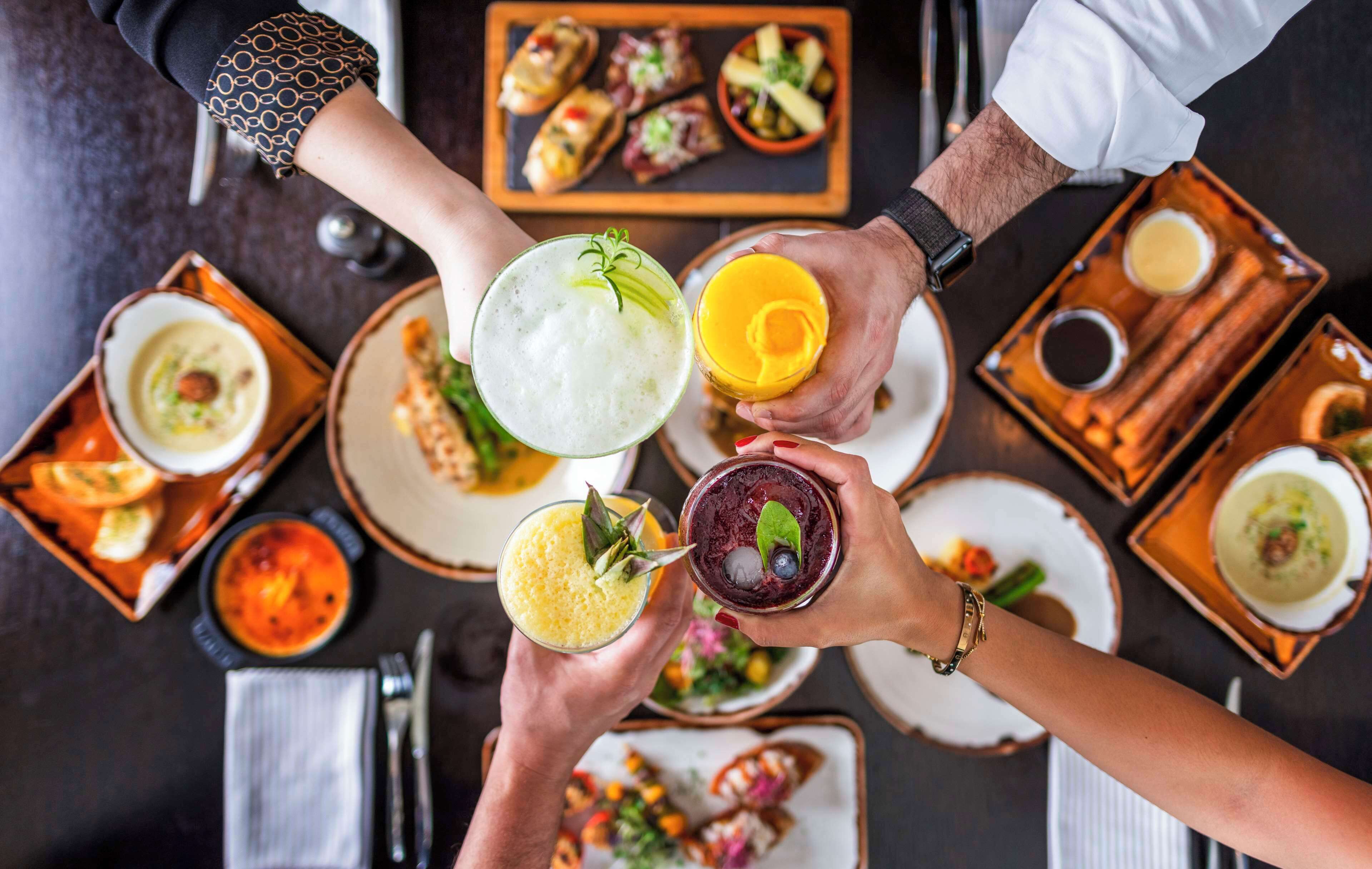 Um grupo de pessoas segurando bebidas em uma mesa de jantar com diversos pratos e uma bandeja de aperitivos.