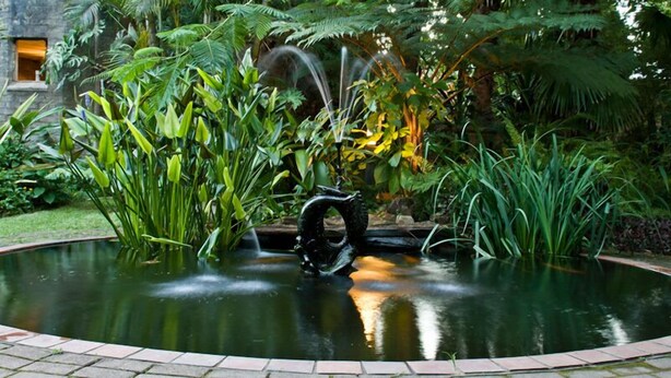 Um lago circular com uma escultura central, rodeado por vegetação exuberante e um edifício de pedra ao fundo.