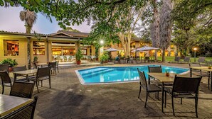 Uma área de piscina ao ar livre com mesas e cadeiras, cercada por vegetação e um edifício com várias varandas.