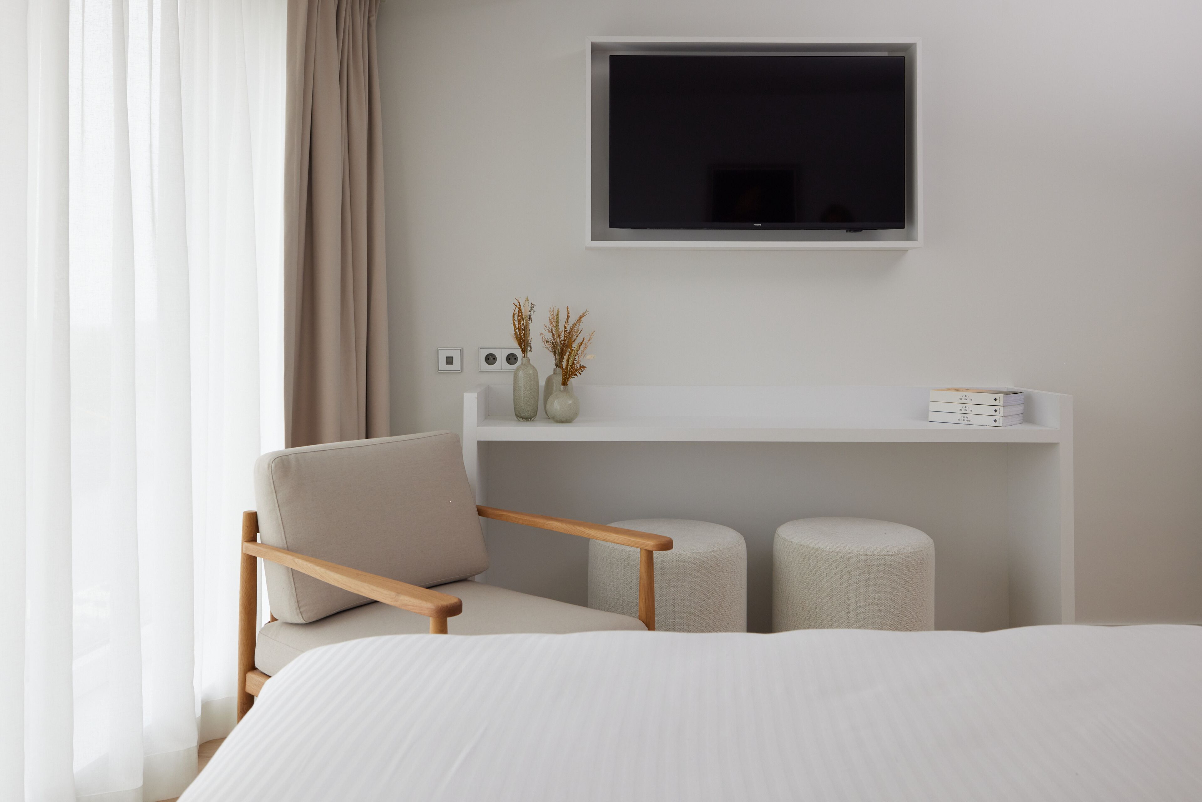 Quarto de hotel moderno com uma cama, uma cadeira, uma TV fixada na parede e uma pequena prateleira com objetos decorativos.