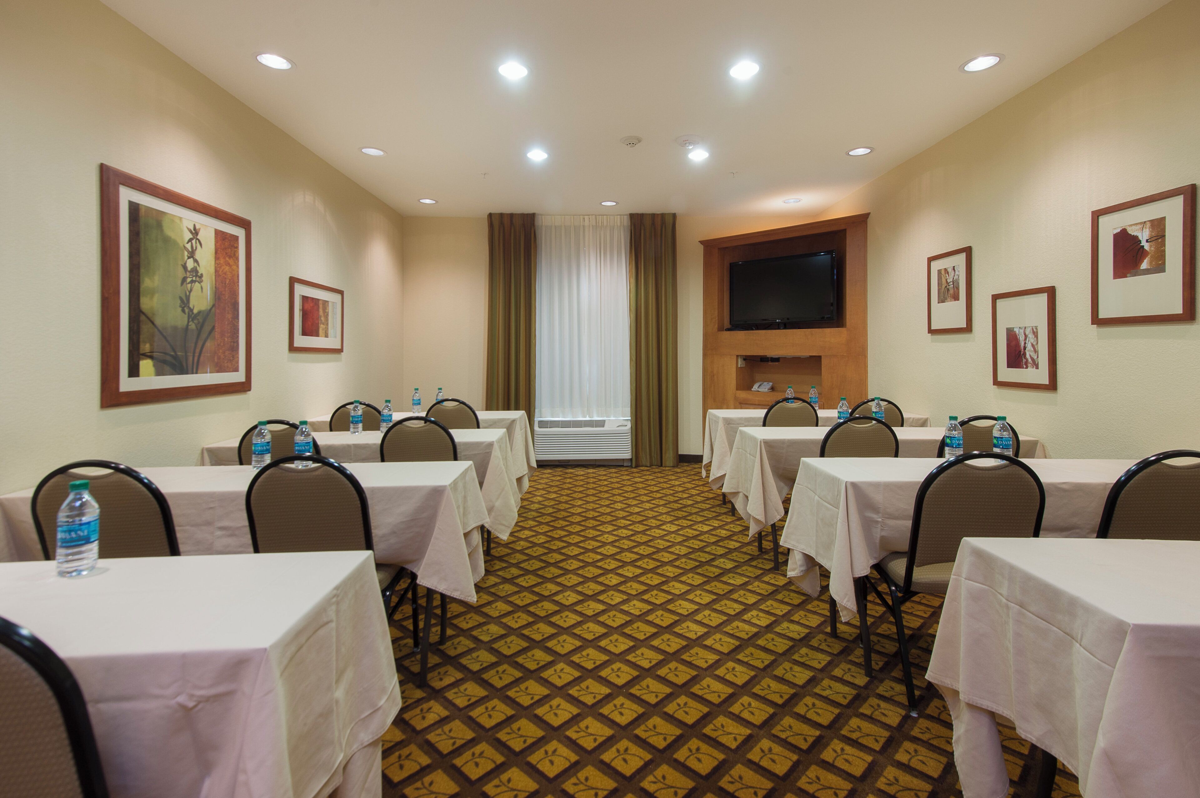Uma sala de conferências de hotel com mesas, cadeiras, uma televisão e obras de arte emolduradas nas paredes.
