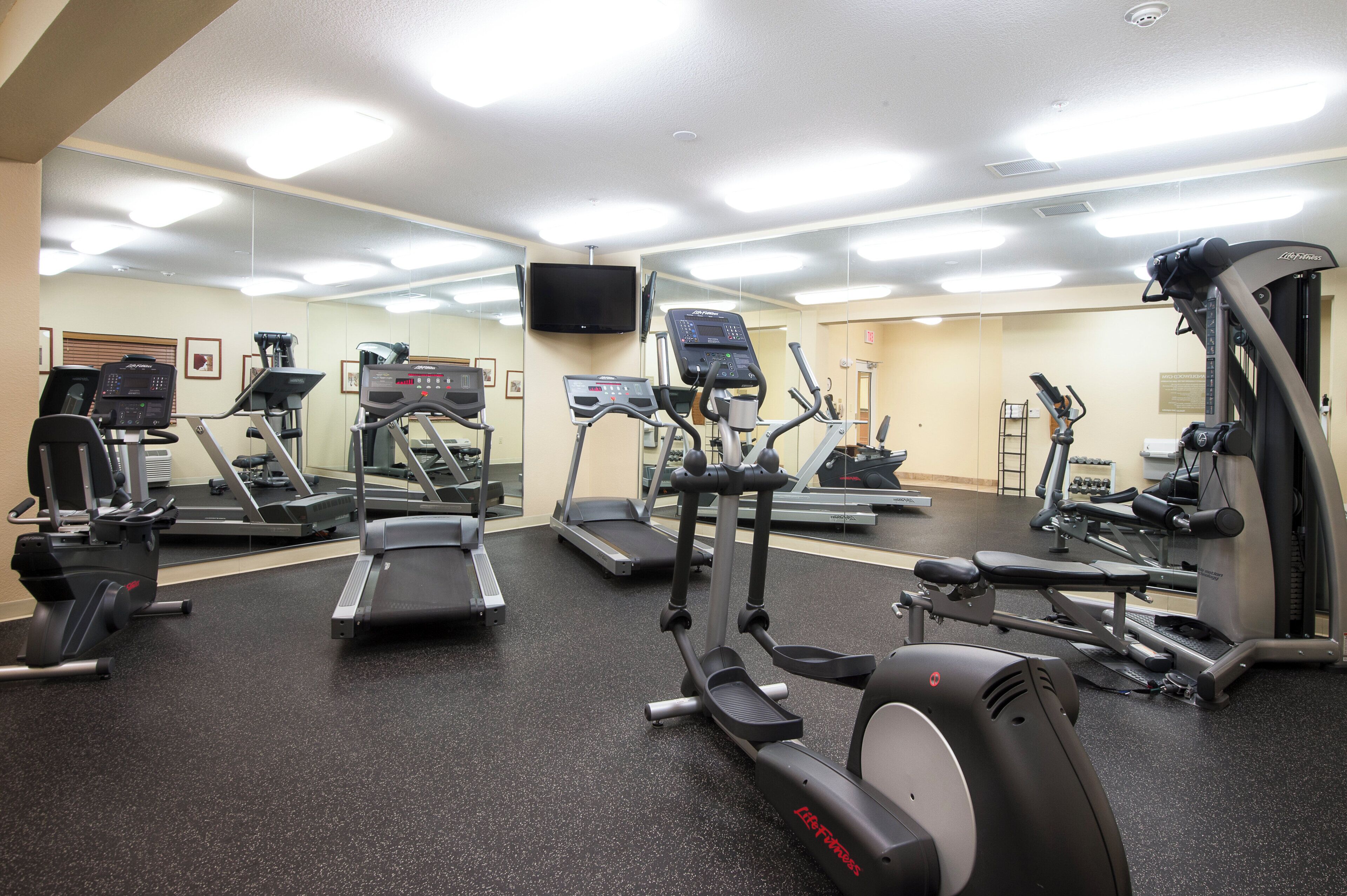 Academia com diversos equipamentos de exercício, incluindo esteiras e elípticos, e uma televisão presa à parede.