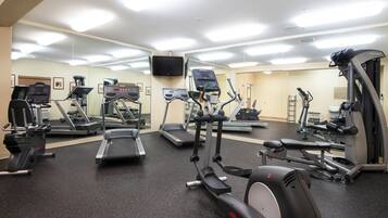 Academia com diversos equipamentos de exercício, incluindo esteiras e elípticos, e uma televisão presa à parede.