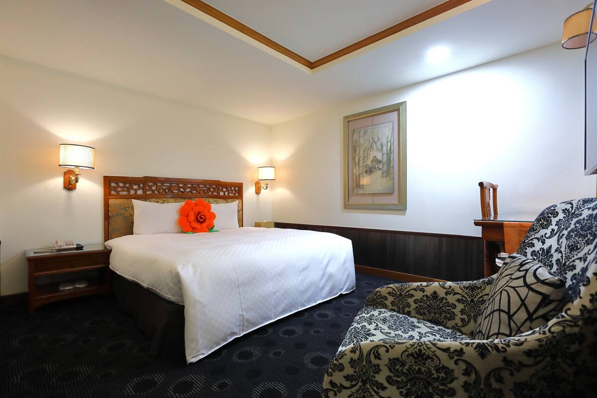 Quarto de hotel com cama, duas luminárias de cabeceira, mesa de apoio, uma cadeira e um quadro na parede.