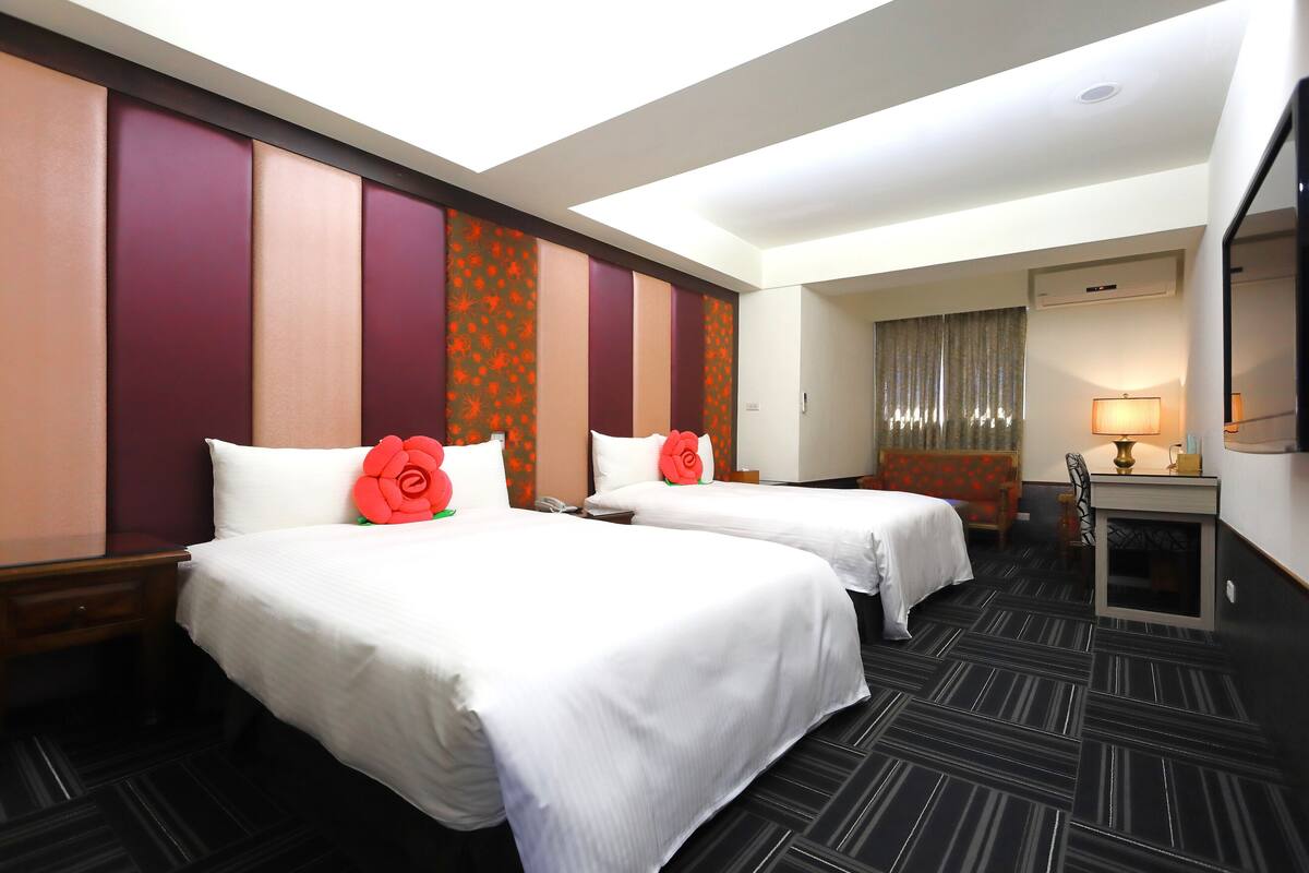 Quarto de hotel com duas camas, uma escrivaninha, uma cadeira, um abajur e um espelho.