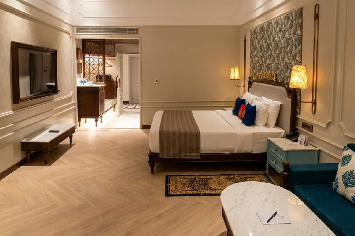 Quarto de hotel com cama, mesa de cabeceira, luminária, televisão e escrivaninha com cadeira.
