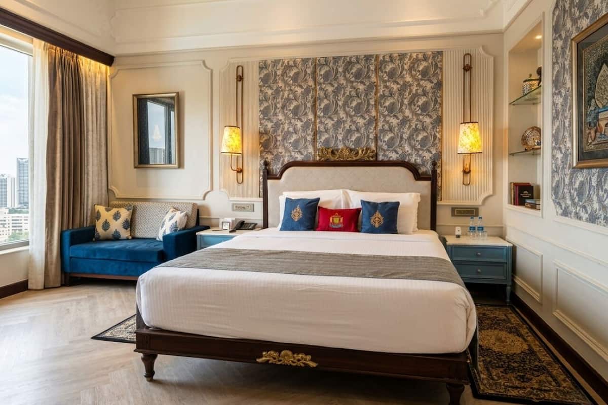 Quarto de hotel com uma cama grande, um sofá, uma mesa de cabeceira, uma luminária, um espelho e vista para a cidade.