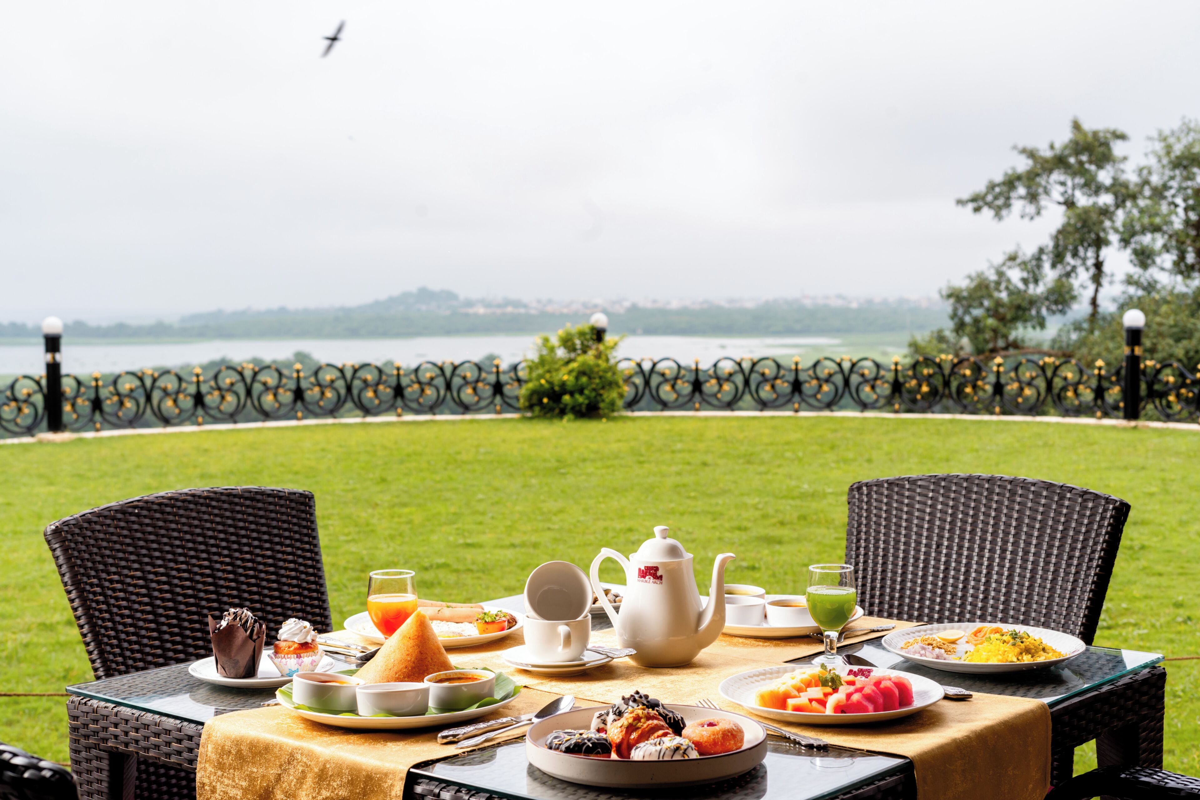 Mesa de jantar ao ar livre com itens de café da manhã, cadeiras e vista para um corpo d'água.