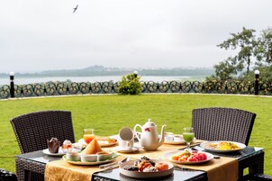 Mesa de jantar ao ar livre com itens de café da manhã, cadeiras e vista para um corpo d'água.
