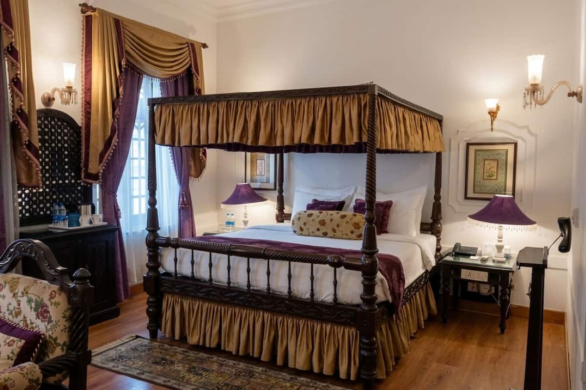 Uma cama com dossel e cortina, uma mesa de cabeceira com abajur, uma cadeira com estofamento floral e um tapete estampado sobre um piso de madeira.