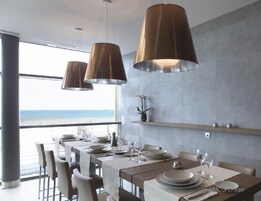 Uma sala de jantar moderna com vista para o oceano, apresentando uma mesa comprida posta para refeições, luminárias pendentes e uma decoração minimalista.