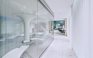 Um interior moderno com paredes de vidro, mobiliário branco e uma escada que leva a um andar superior.