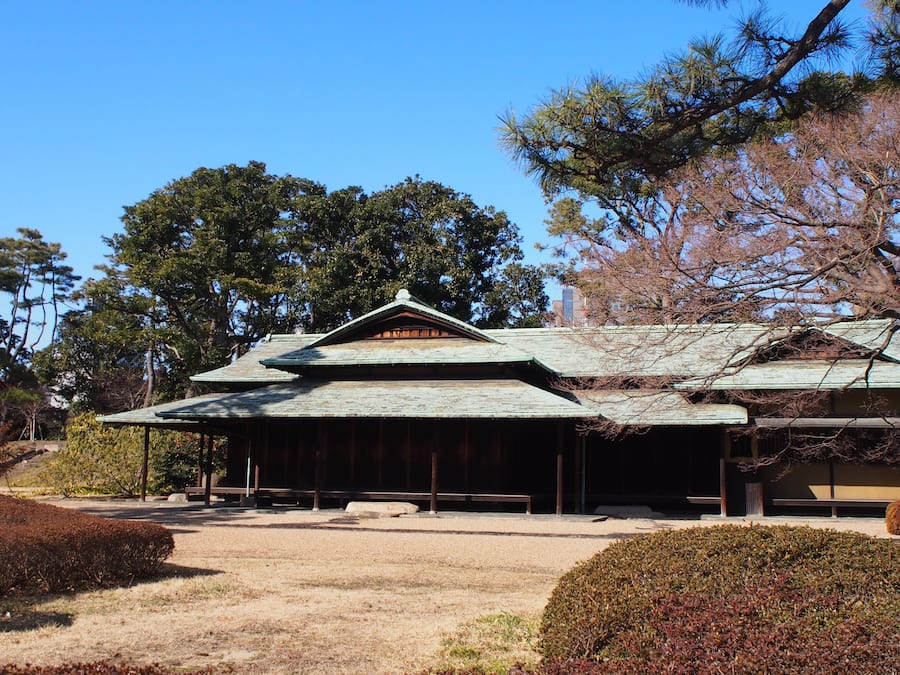 Um edifício japonês tradicional com telhado em telhas, rodeado por arbustos e árvores bem cuidados.
