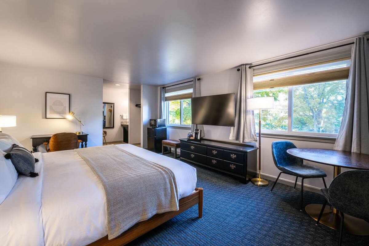 Quarto de hotel com uma cama grande, uma escrivaninha, uma cadeira, televisão e janela com cortinas.