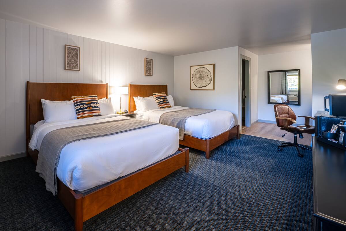 Quarto de hotel com duas camas de madeira, uma escrivaninha com cadeira, um espelho e um quadro na parede.