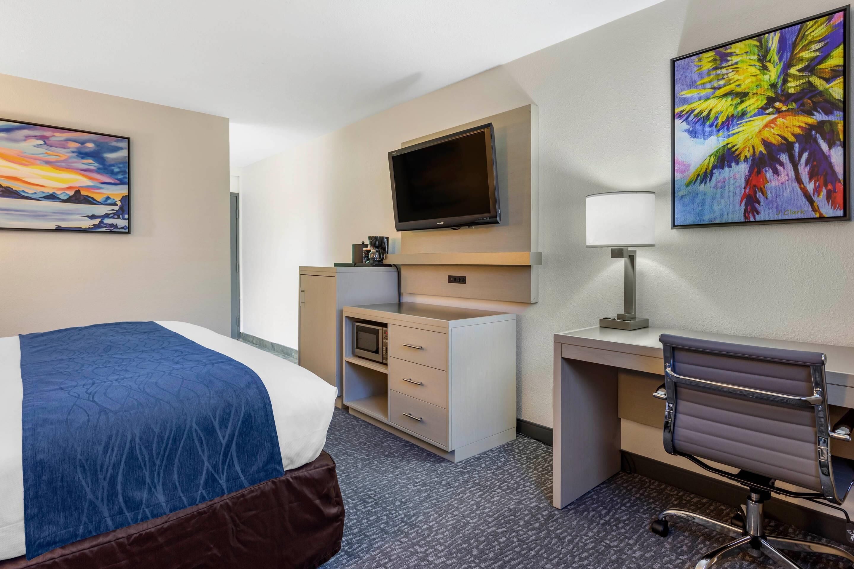 Quarto de hotel com cama, escrivaninha, cadeira, televisão e obras de arte nas paredes.