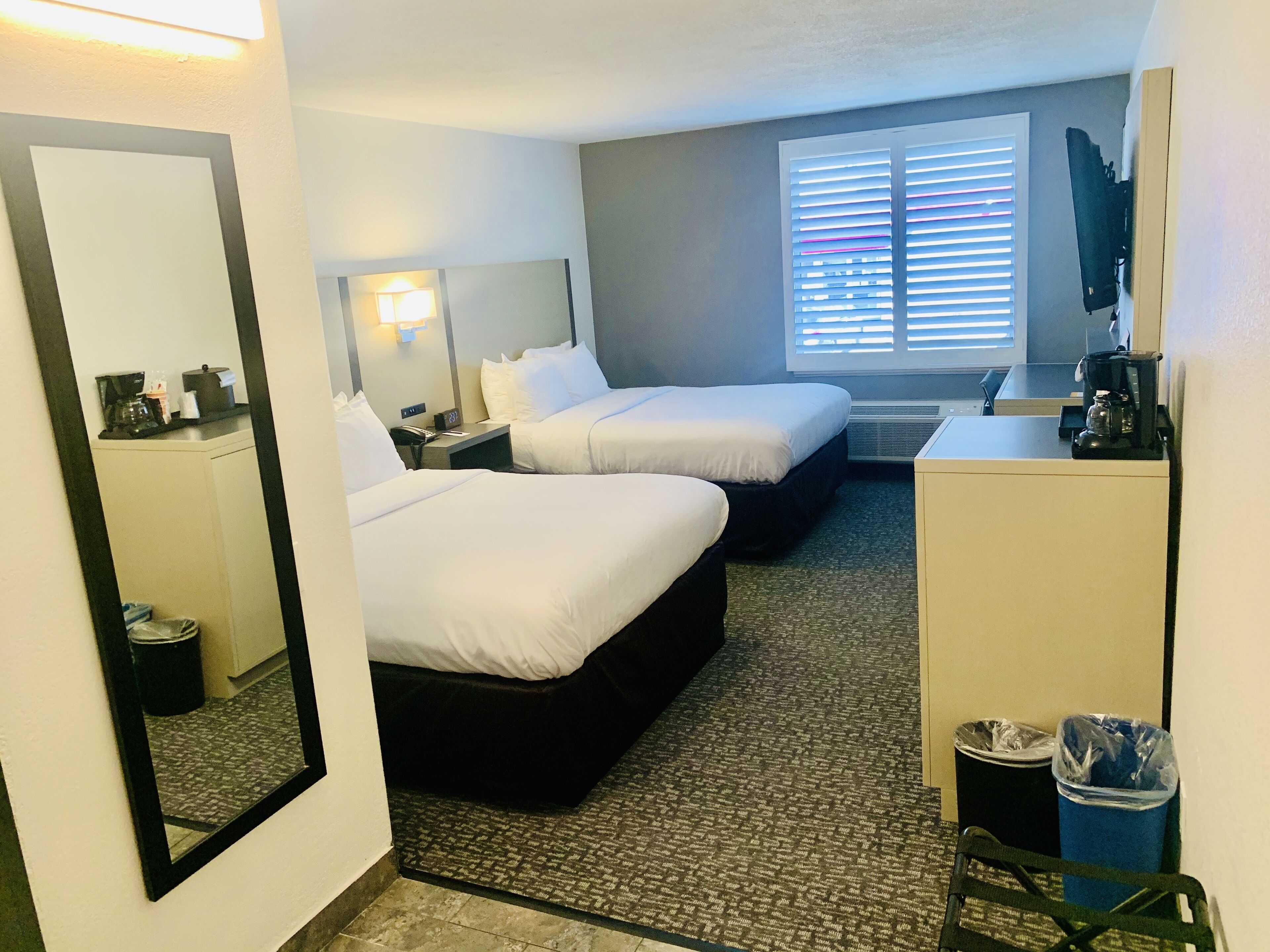 Quarto de hotel com duas camas, uma secretária com máquina de café, televisão na parede e janela com persianas.