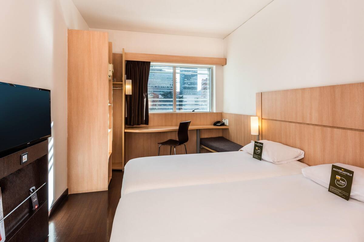 Quarto de hotel com cama, escrivaninha, cadeira, televisão e janela com cortinas.